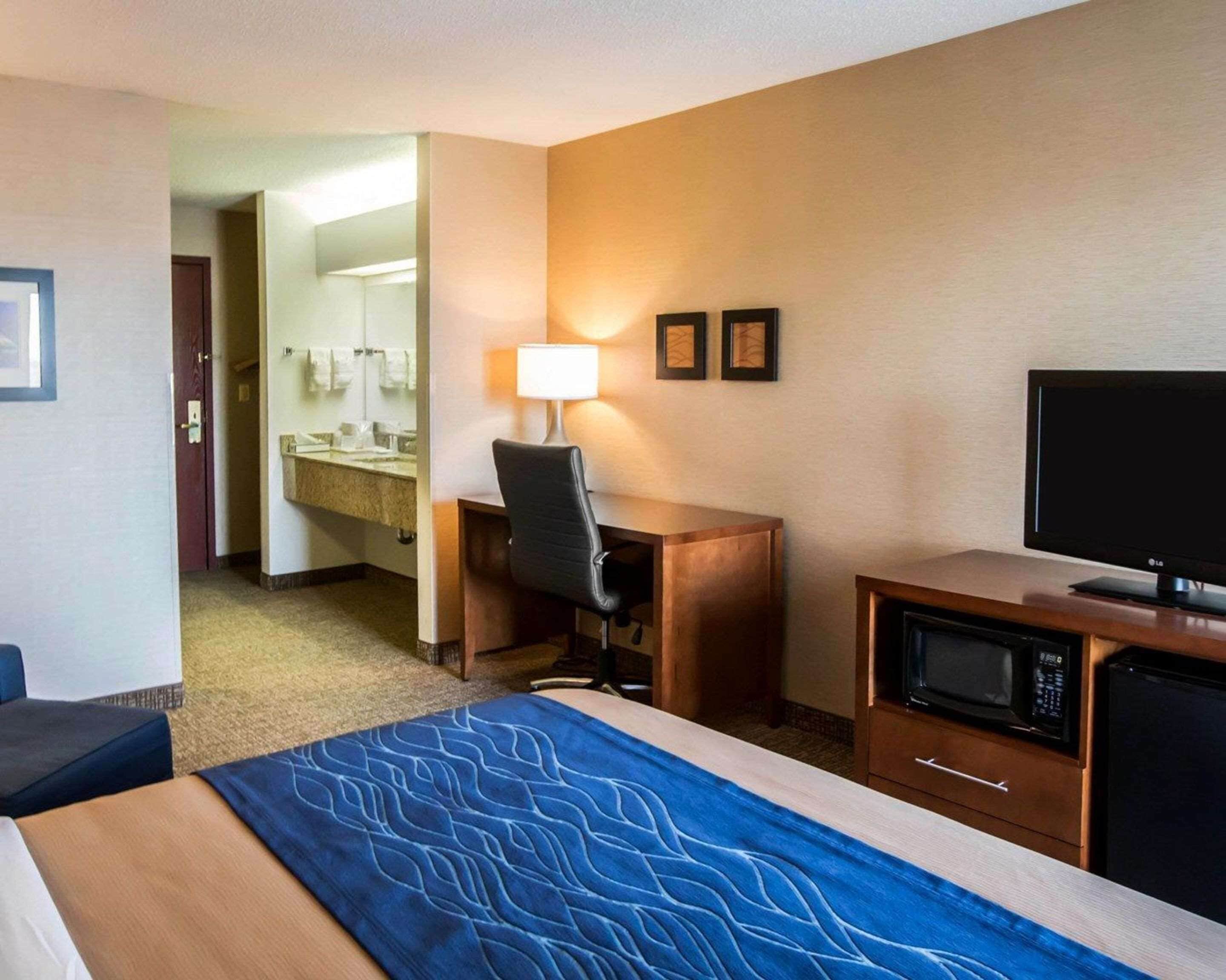 Quarto de hotel com cama, escrivaninha e cadeira, televisão, micro-ondas e banheiro visível por uma porta aberta.