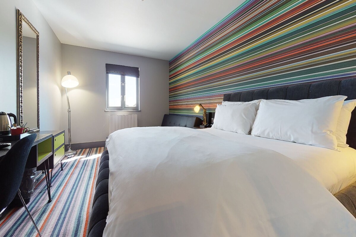 Quarto de hotel com uma cama grande, uma escrivaninha, uma cadeira, um abajur e uma parede colorida listrada.
