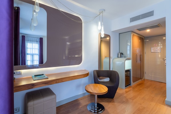 Quarto de hotel moderno com toucador de madeira, um banquinho redondo, um espelho grande e cabine de duche em vidro.