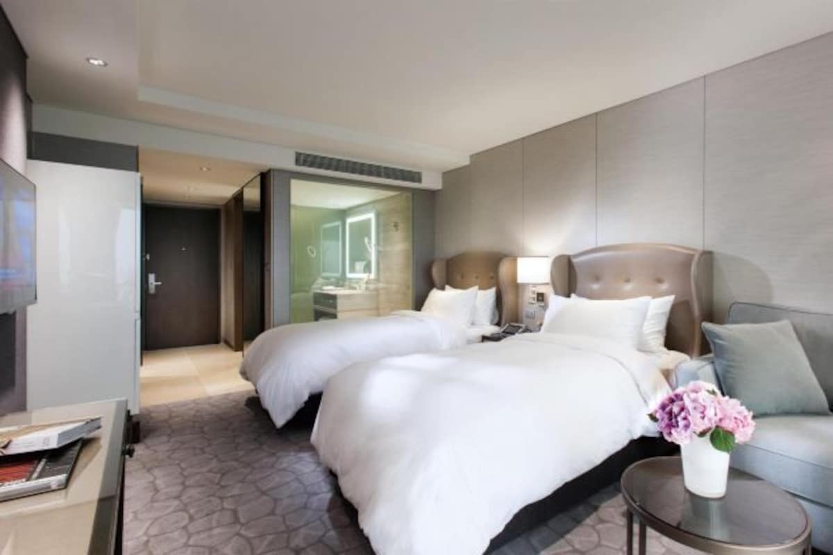 Quarto de hotel com duas camas, um sofá, uma TV e uma pequena mesa com um vaso de flores.