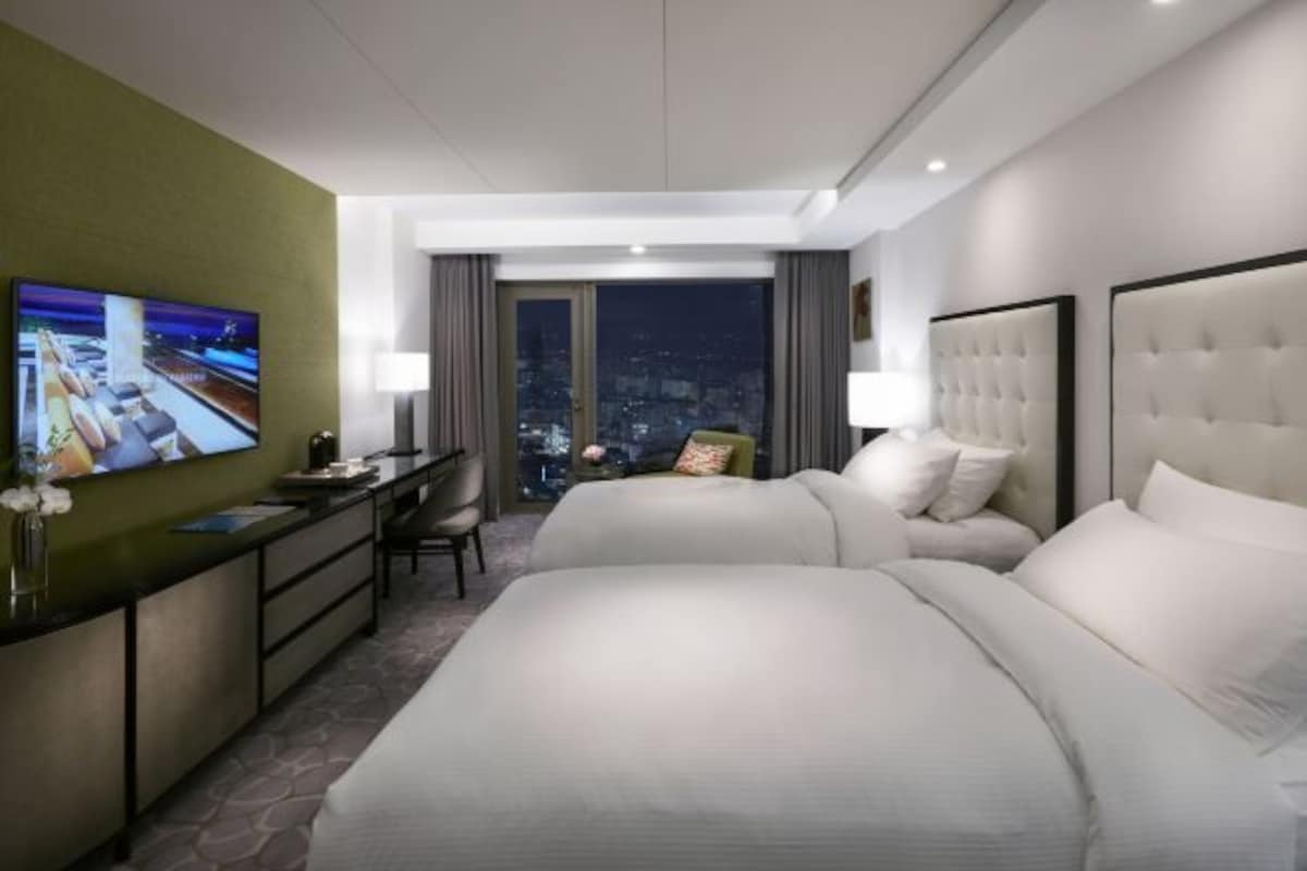 Quarto de hotel com duas camas, uma escrivaninha, uma cadeira, uma televisão e vista para a cidade.