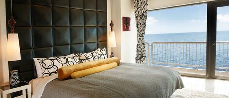 Quarto com uma cama grande, cabeceira, mesa de cabeceira, luminária e vista para o mar através de uma varanda.