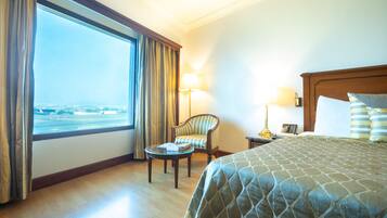 Quarto de hotel com uma janela grande, uma cama, uma cadeira, uma mesa pequena e uma mesinha de cabeceira com abajur.