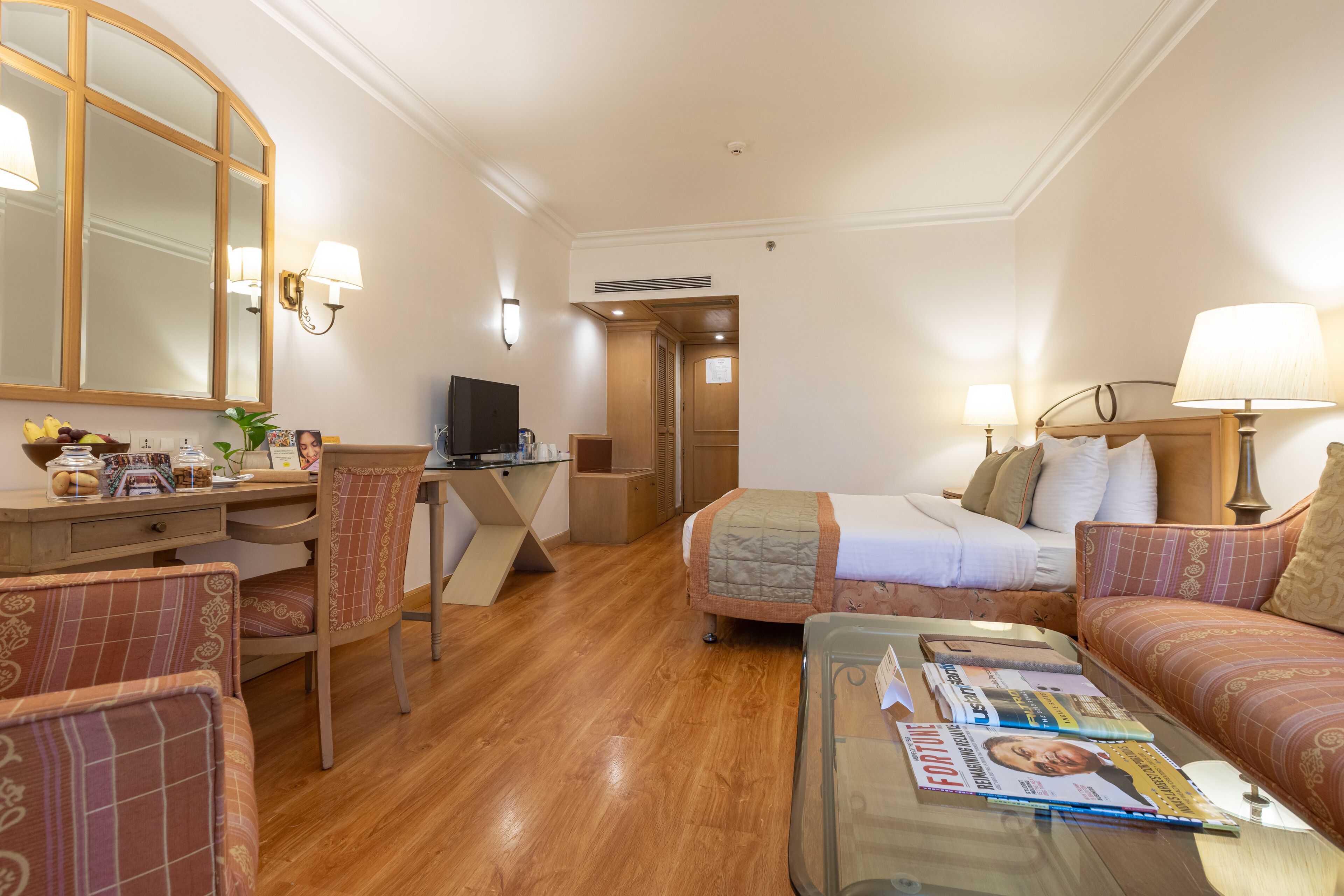 Quarto de hotel com cama, escrivaninha com computador, cadeira, sofá e revista na mesa de centro.