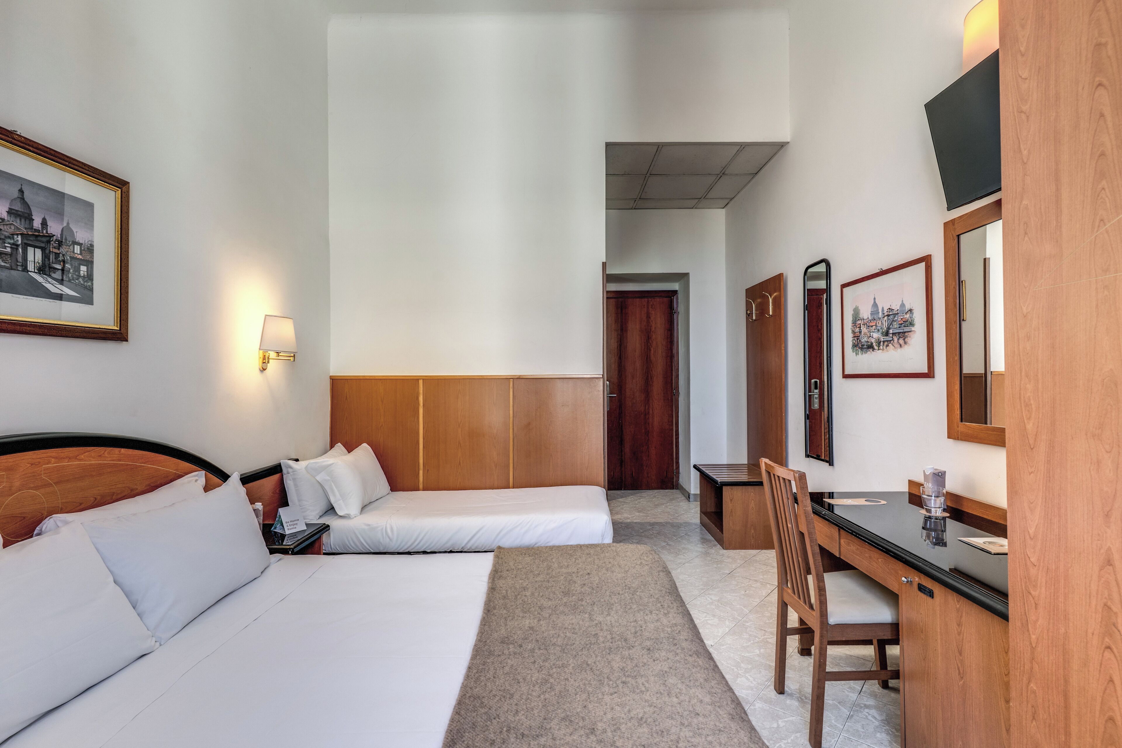 Quarto de hotel com duas camas, uma escrivaninha, uma cadeira, uma luminária, um espelho e um quadro na parede.