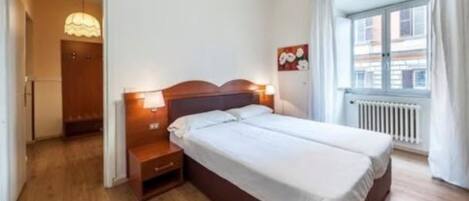 Uma cama de casal com cabeceira de madeira, uma mesinha de cabeceira com luminária, um radiador e uma janela com cortinas.