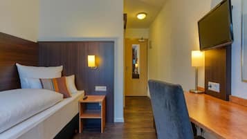 Quarto de hotel com cama, escrivaninha, cadeira, televisão e corredor que dá para outro quarto.