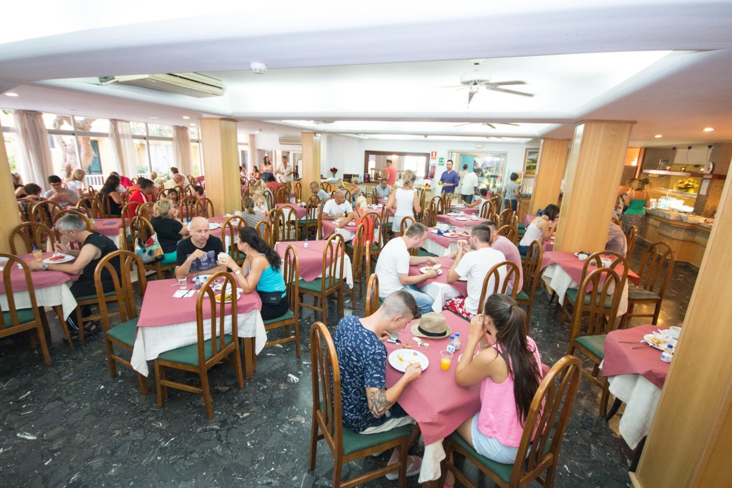Uma sala de jantar com várias mesas e cadeiras, janelas amplas e um ventilador de teto.