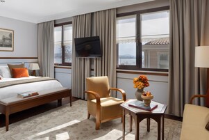 Quarto de hotel com cama, televisão na parede, uma cadeira, uma mesa pequena com um vaso de flores e vista para um edifício pela janela.