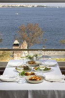 Uma mesa posta com diversos pratos e copos de vinho branco, com vista para um corpo d'água e edifícios ao longe, além de uma árvore.