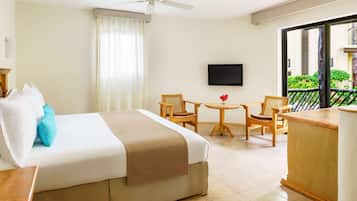 Quarto de hotel com uma cama, uma escrivaninha, uma cadeira, uma mesinha, um ventilador de teto e uma televisão na parede.