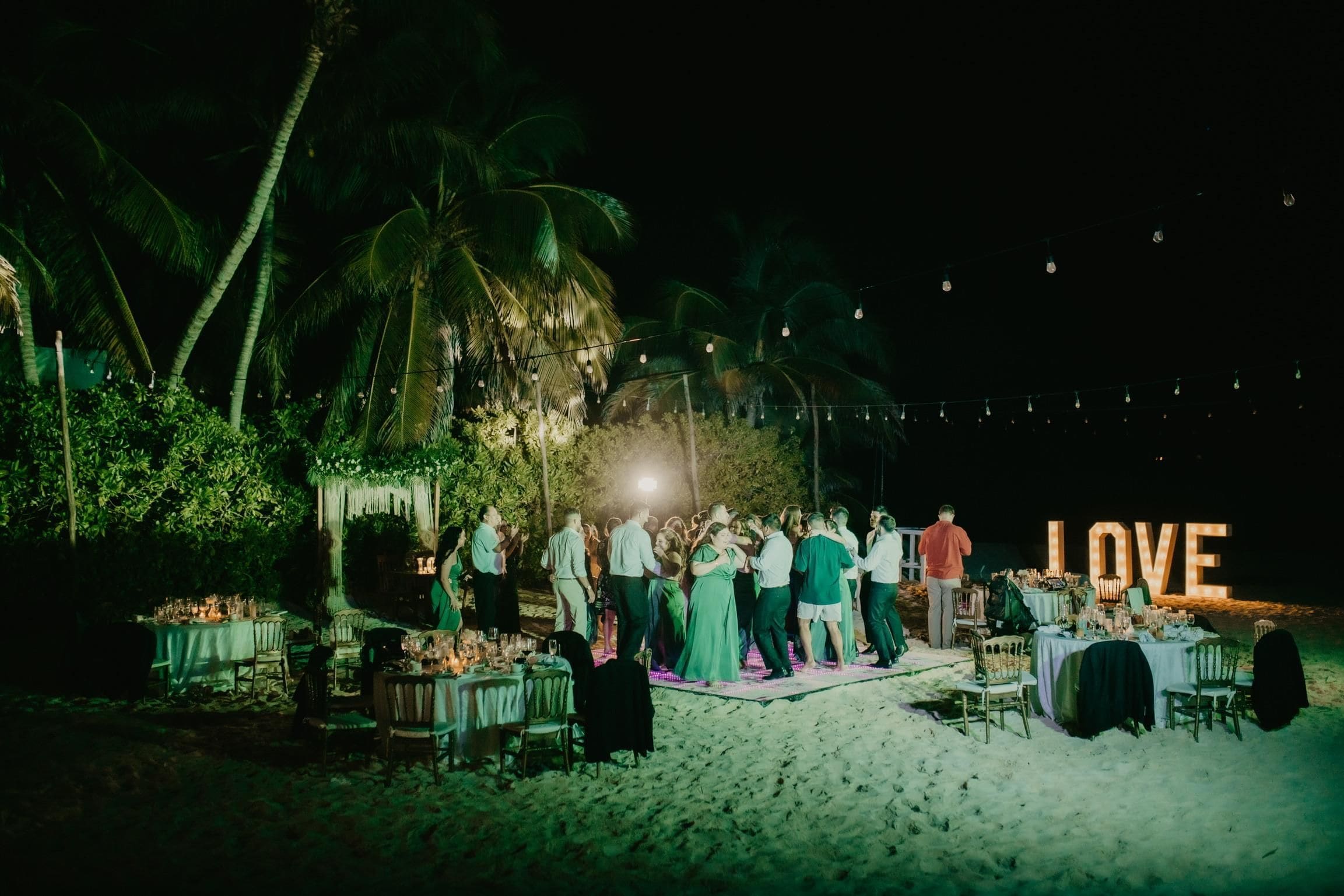Evento ao ar livre com mesas, cadeiras, palmeiras e um grande letreiro iluminado 'LOVE'.