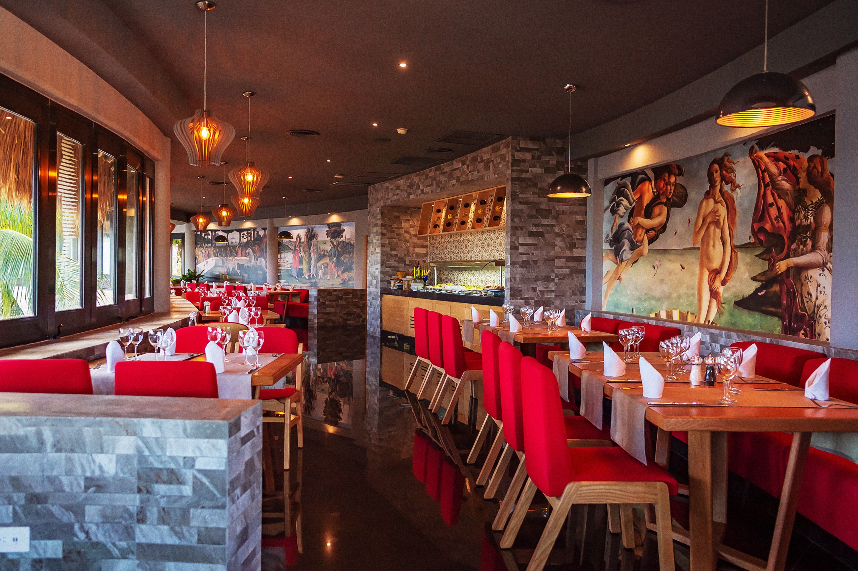 Restaurante com assentos estofados em vermelho, parede com detalhes em pedra e um mural.