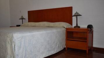 Quarto com cabeceira de madeira, cama com colcha estampada, mesa de cabeceira com abajur e um pequeno armário de madeira.