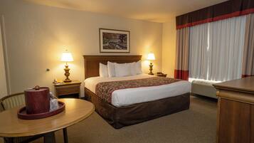 Quarto de hotel com uma cama, duas luminárias de cabeceira, uma mesa com uma cadeira, um quadro na parede e uma janela com cortinas.