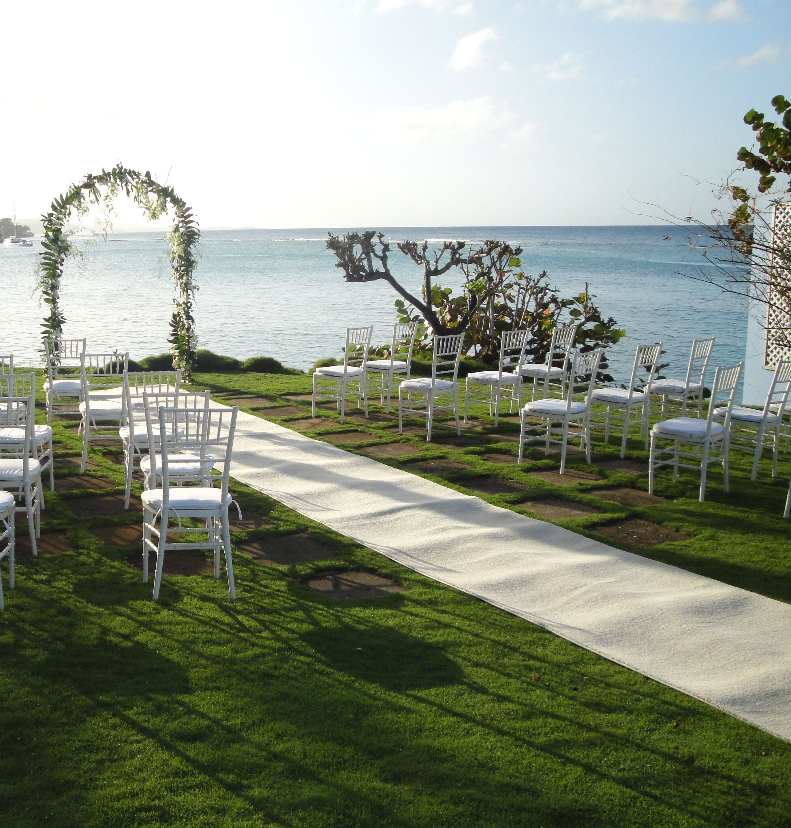 Passarela de casamento com cadeiras brancas e um tapete branco que leva a um arco decorado à beira-mar.