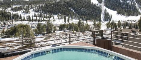 Uma banheira de hidromassagem com vista para uma paisagem montanhosa coberta de neve, com área de piscina e um edifício ao primeiro plano.