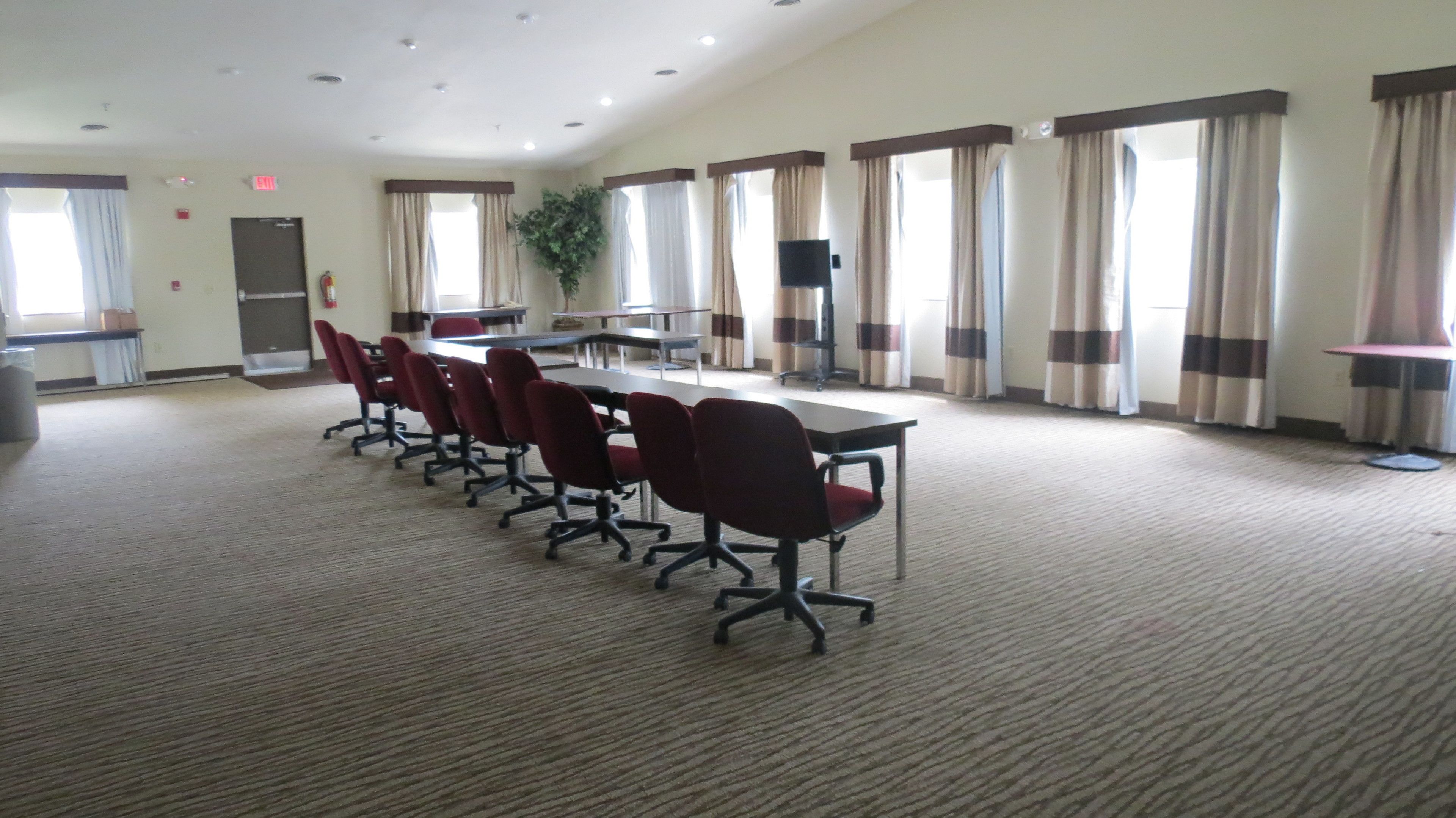 Uma sala de conferências com uma mesa comprida e cadeiras vermelhas, grandes janelas com cortinas e uma planta.
