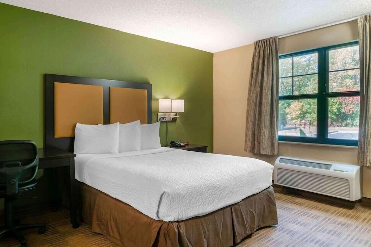 Quarto de hotel com cama, escrivaninha, cadeira, luminária, ar condicionado e janela com cortinas.