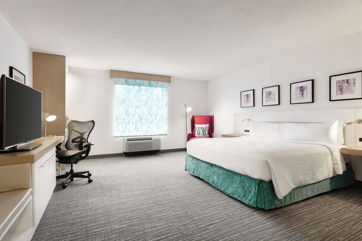 Quarto de hotel com cama, escrivaninha, cadeira, televisão, aparelho de ar condicionado e quadros na parede.
