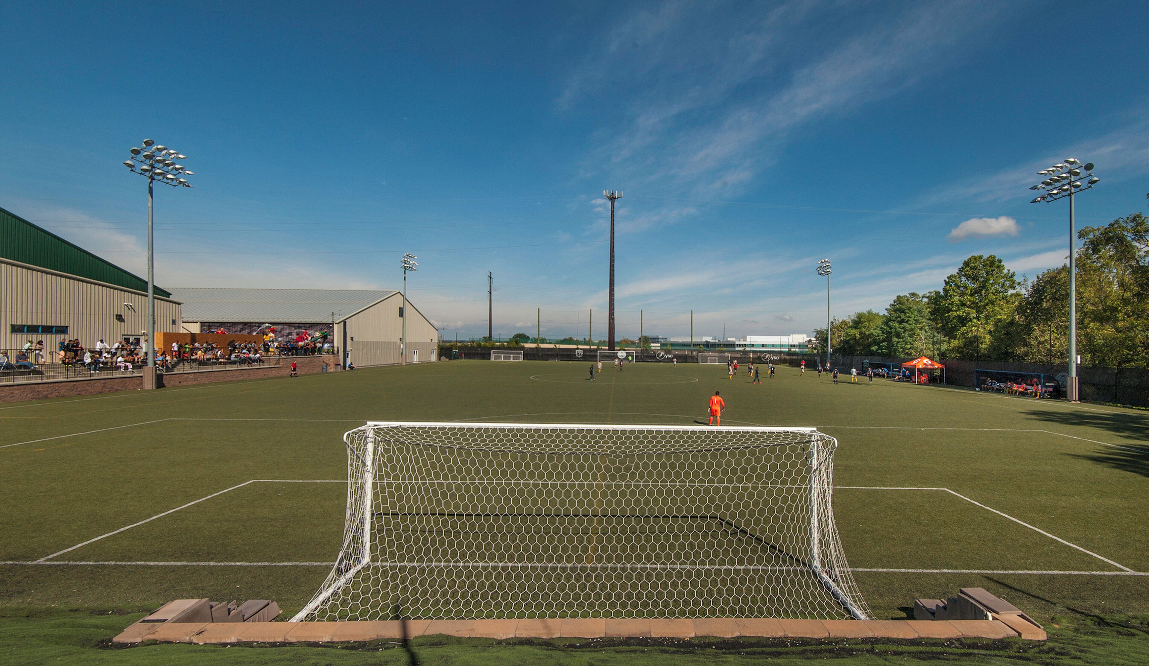 Um campo de futebol com um gol, refletores e um edifício grande ao fundo.