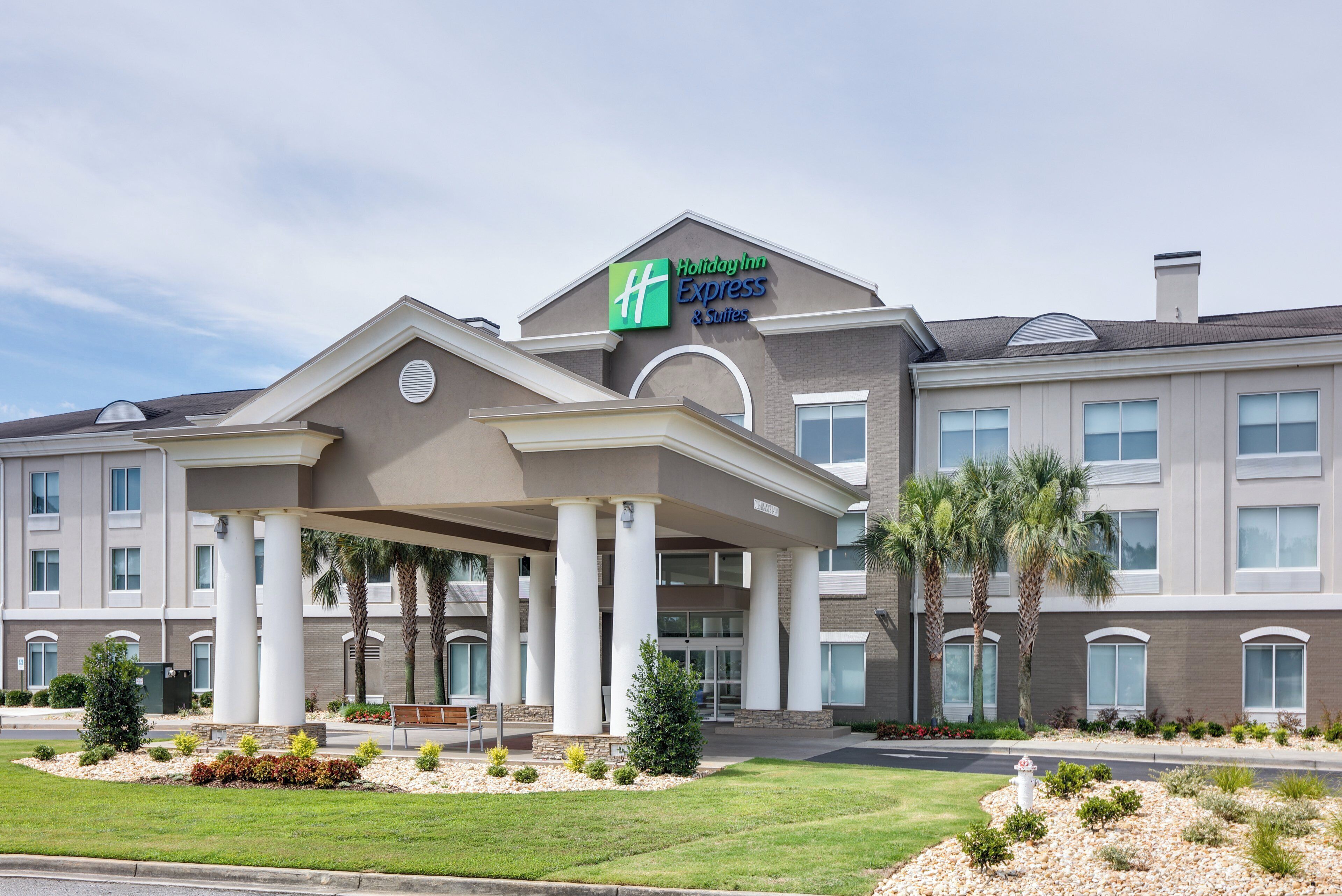 Um hotel Holiday Inn Express & Suites com entrada coberta, vários andares e um jardim frontal ajardinado.