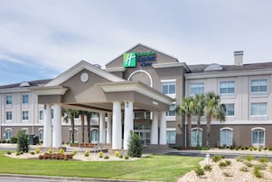Um hotel Holiday Inn Express & Suites com entrada coberta, vários andares e um jardim frontal ajardinado.