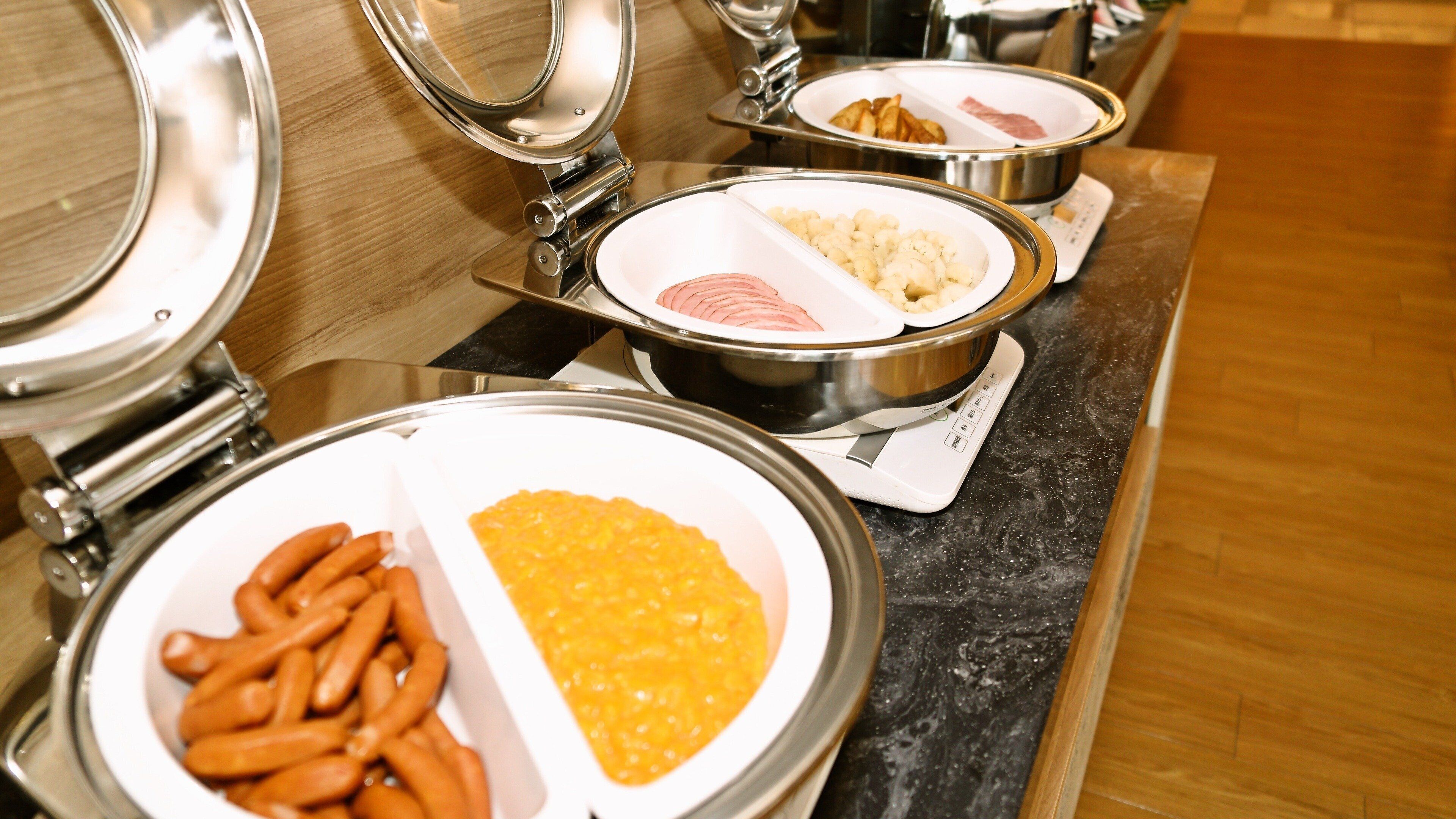 Buffet com diversos pratos, incluindo salsichas, arroz e um prato amarelo, provavelmente um tipo de salada ou gratinado.