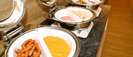 Buffet com diversos pratos, incluindo salsichas, arroz e um prato amarelo, provavelmente um tipo de salada ou gratinado.