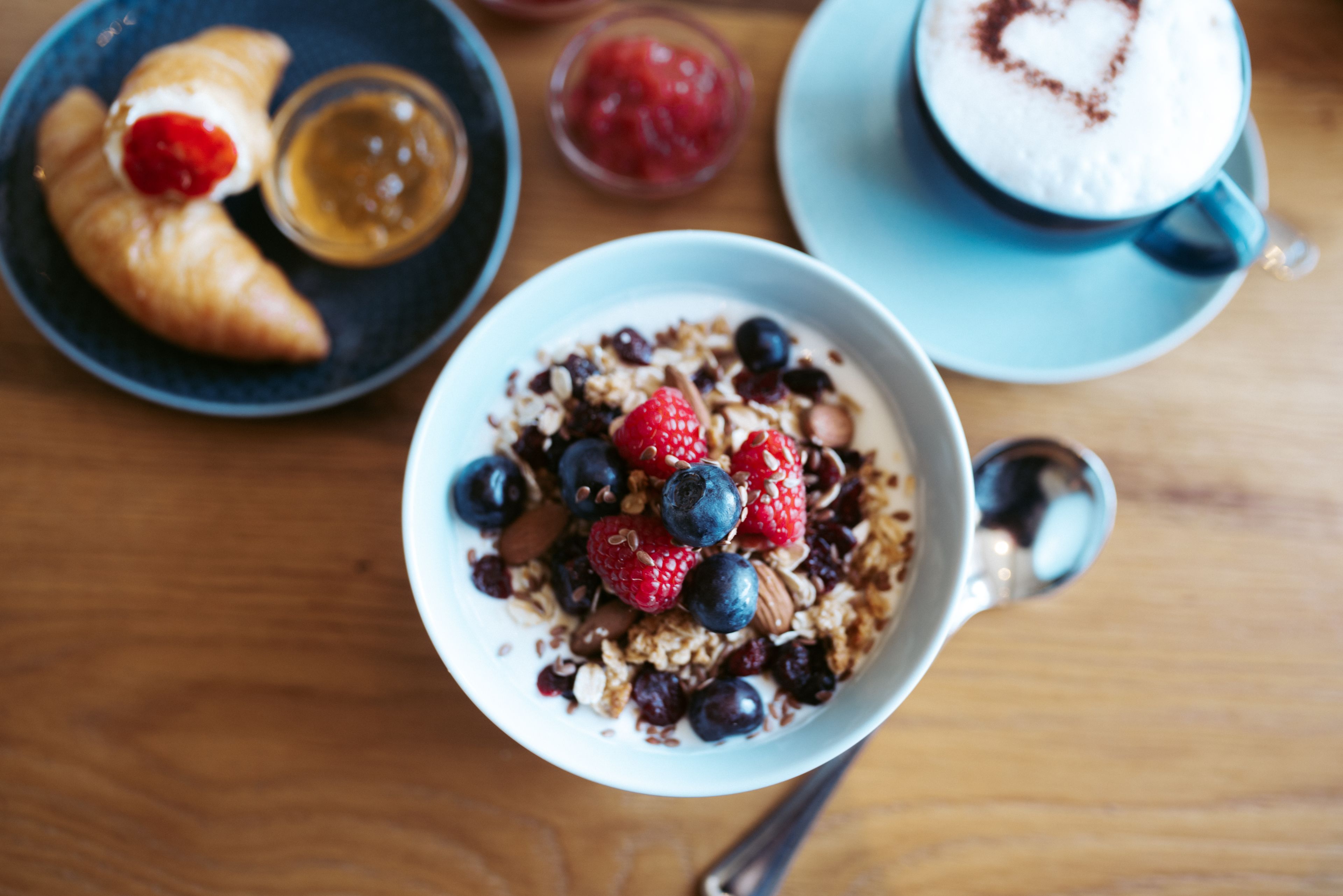 Disposição para o café da manhã com uma tigela de cereal coberta com frutas vermelhas e nozes, um croissant e uma xícara de café com espuma em formato de coração.