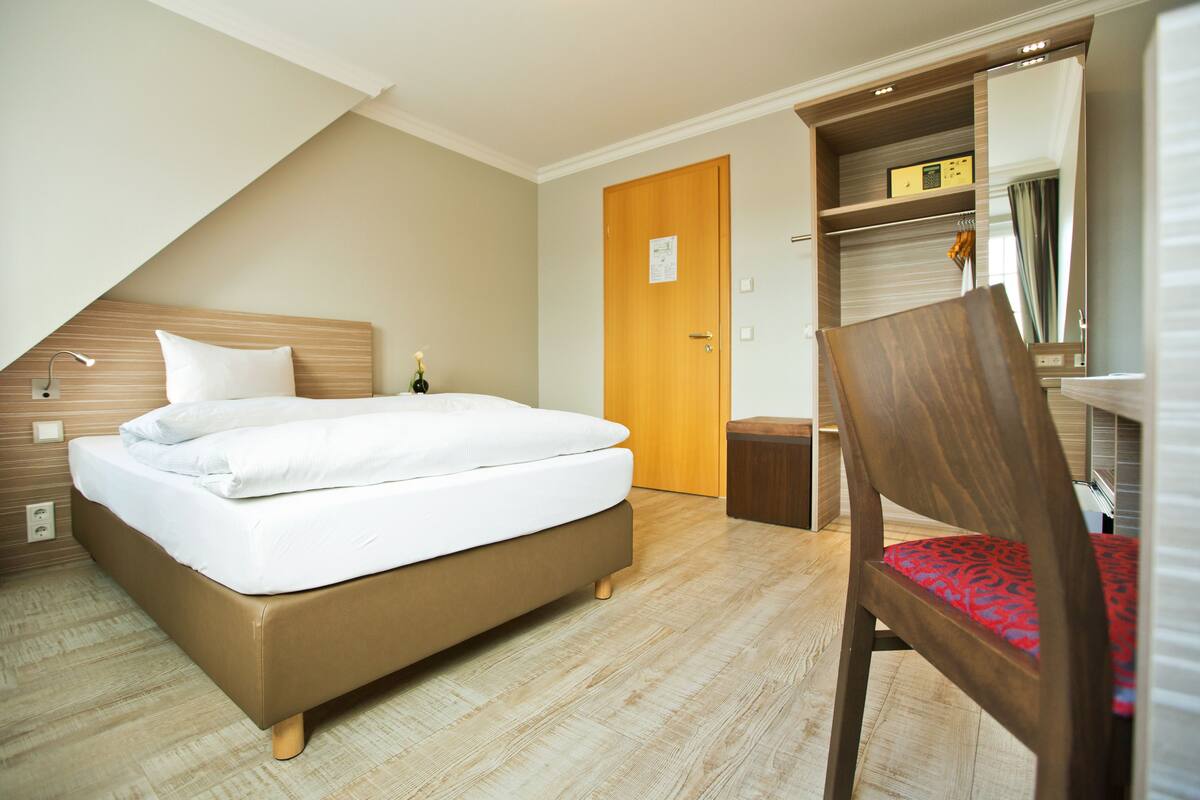 Quarto de hotel com uma cama, uma cadeira de madeira, uma mesa pequena e uma porta.
