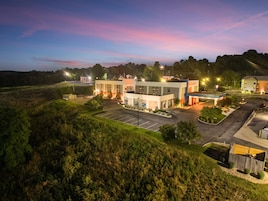 Um hotel moderno com estacionamento, cercado por vegetação, e iluminado por postes de luz.