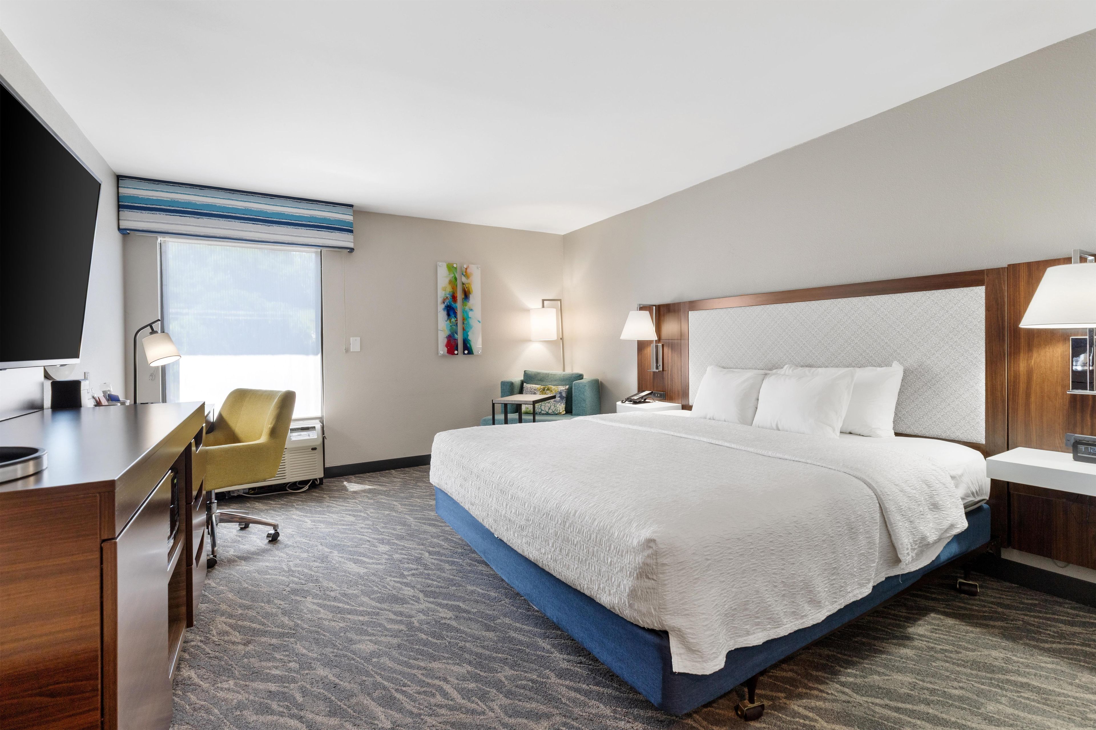 Quarto de hotel com uma cama grande, uma escrivaninha, uma cadeira, uma televisão e uma janela com persianas.