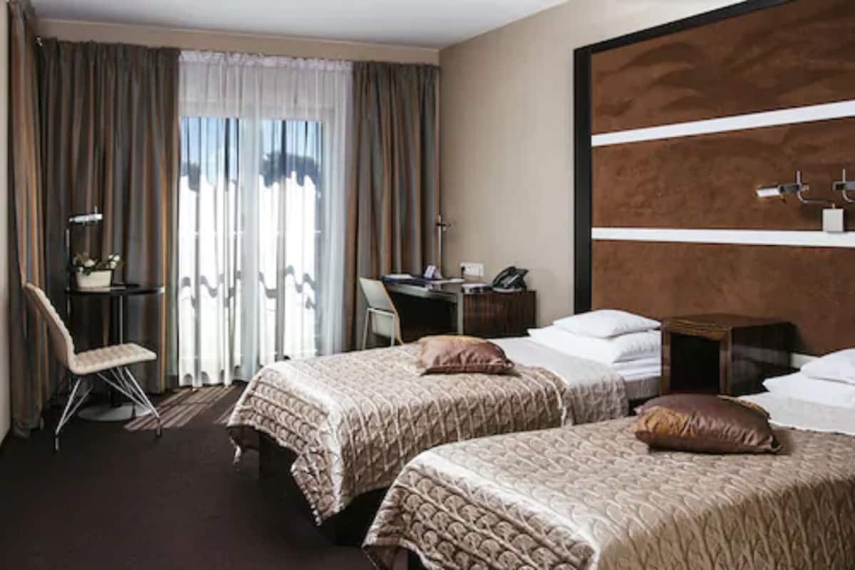 Quarto de hotel com duas camas, uma mesa, uma cadeira e uma janela grande com cortinas.