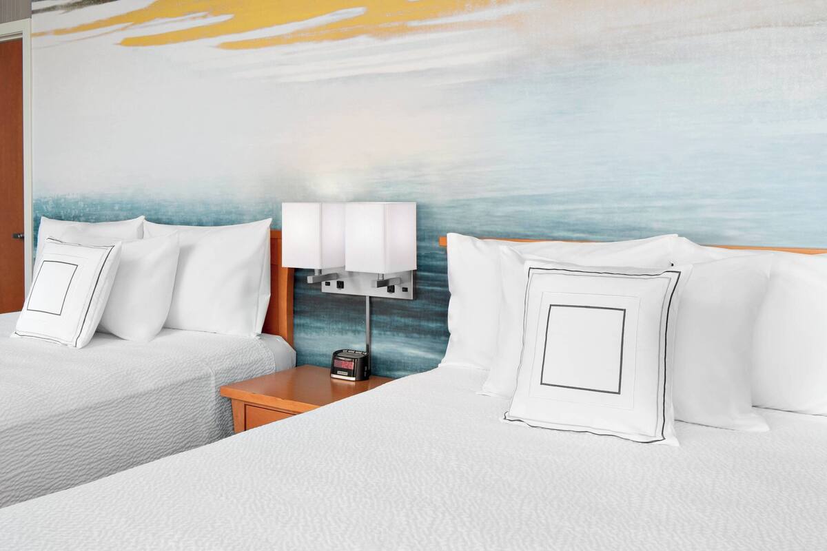 Quarto de hotel com duas camas, uma mesa de cabeceira com abajur e um quadro de paisagem marítima na parede.