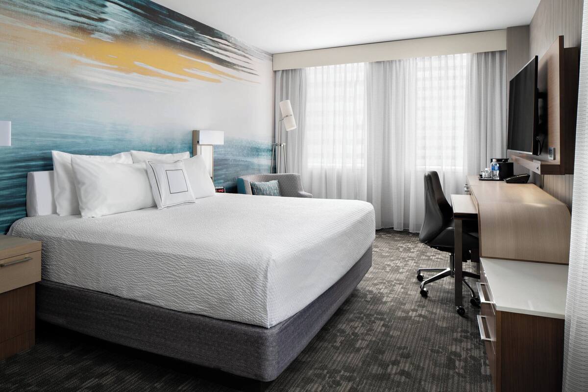 Quarto de hotel com uma cama grande, uma escrivaninha, uma cadeira, uma televisão e um quadro na parede.