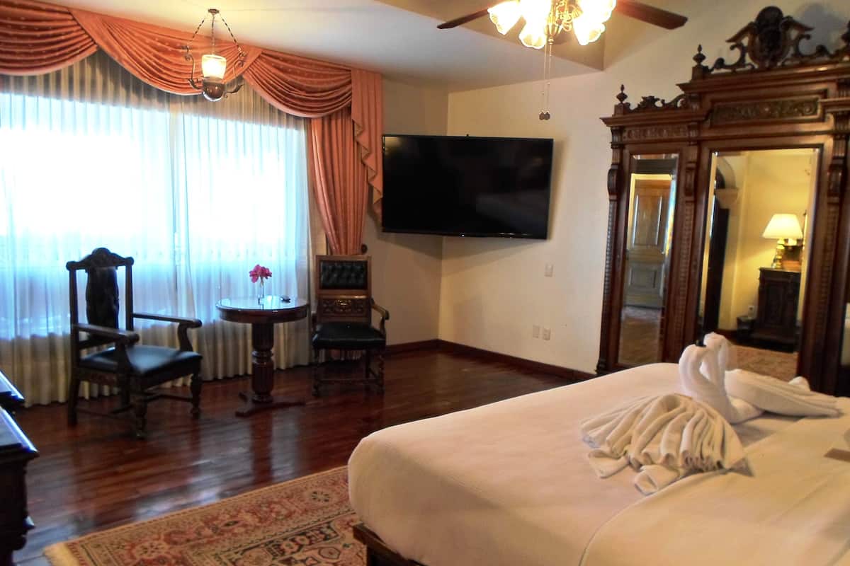 Quarto de hotel com uma cama, televisão, cadeira, mesa pequena e ventoinha no teto.