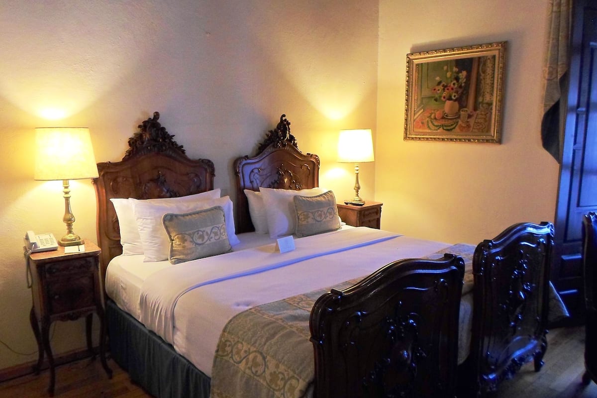 Quarto de hotel com uma cama grande, duas mesas de cabeceira com luminárias, um quadro na parede e uma cadeira.