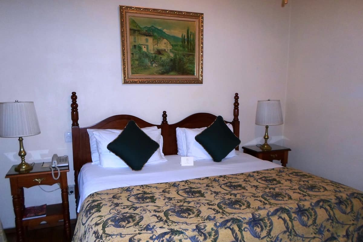 Uma cama bem arrumada com um edredom estampado, duas luminárias de cabeceira, um criado-mudo de madeira com um telefone e um quadro na parede.