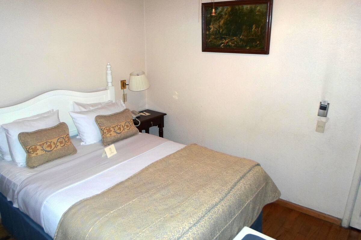 Uma cama bem arrumada com um cobertor bege e duas almofadas decorativas, um abajur e um quadro na parede.