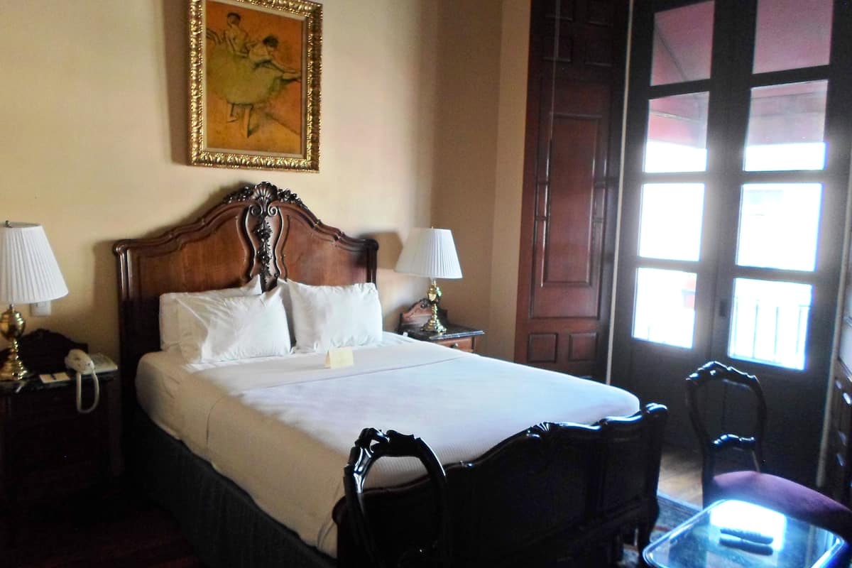 Quarto de hotel com uma cama grande, duas mesas de cabeceira com luminárias, um painel de madeira na cabeceira, um quadro na parede e uma janela com vista.