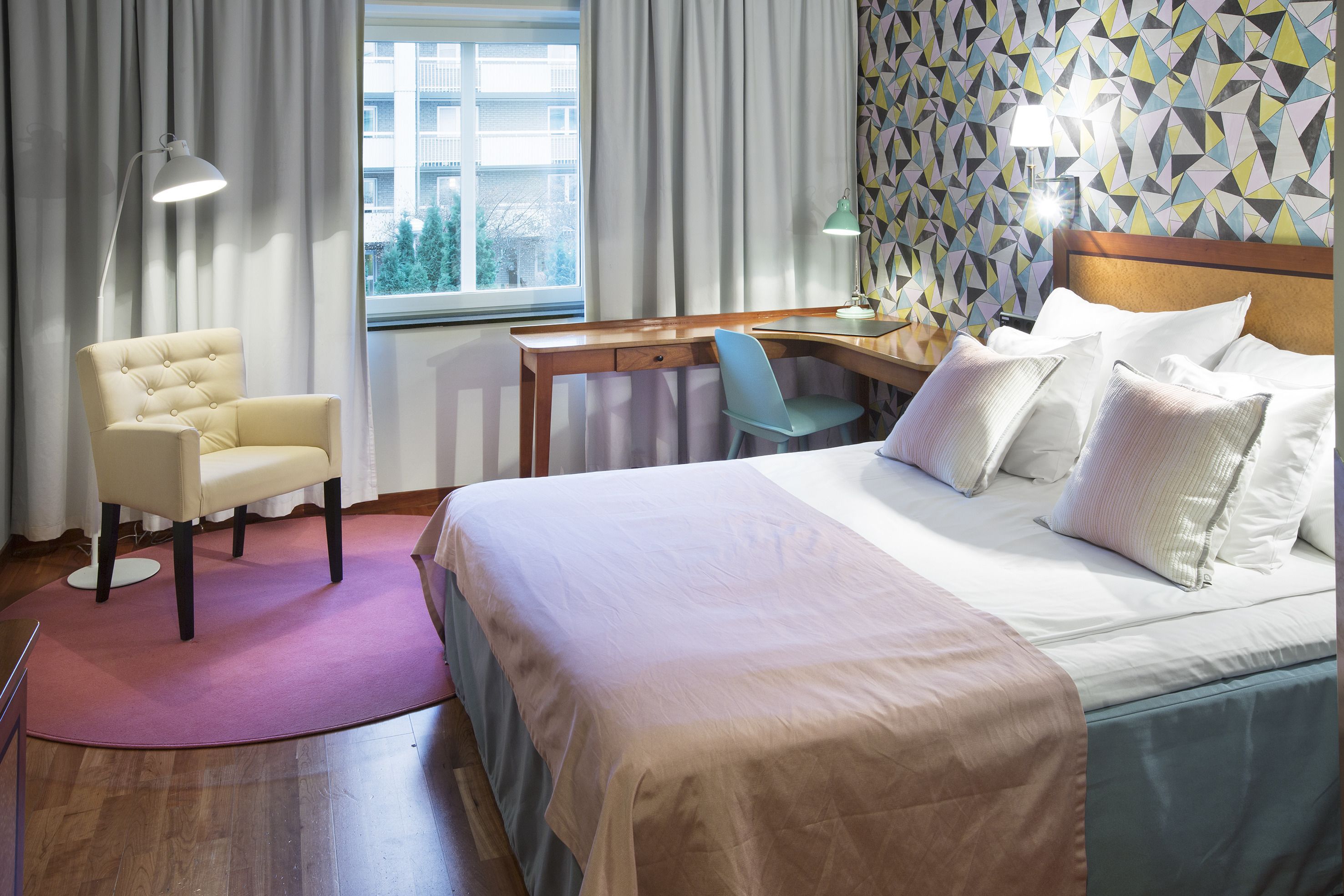 Quarto de hotel com cama, escrivaninha, cadeira e vista para o exterior pela janela.