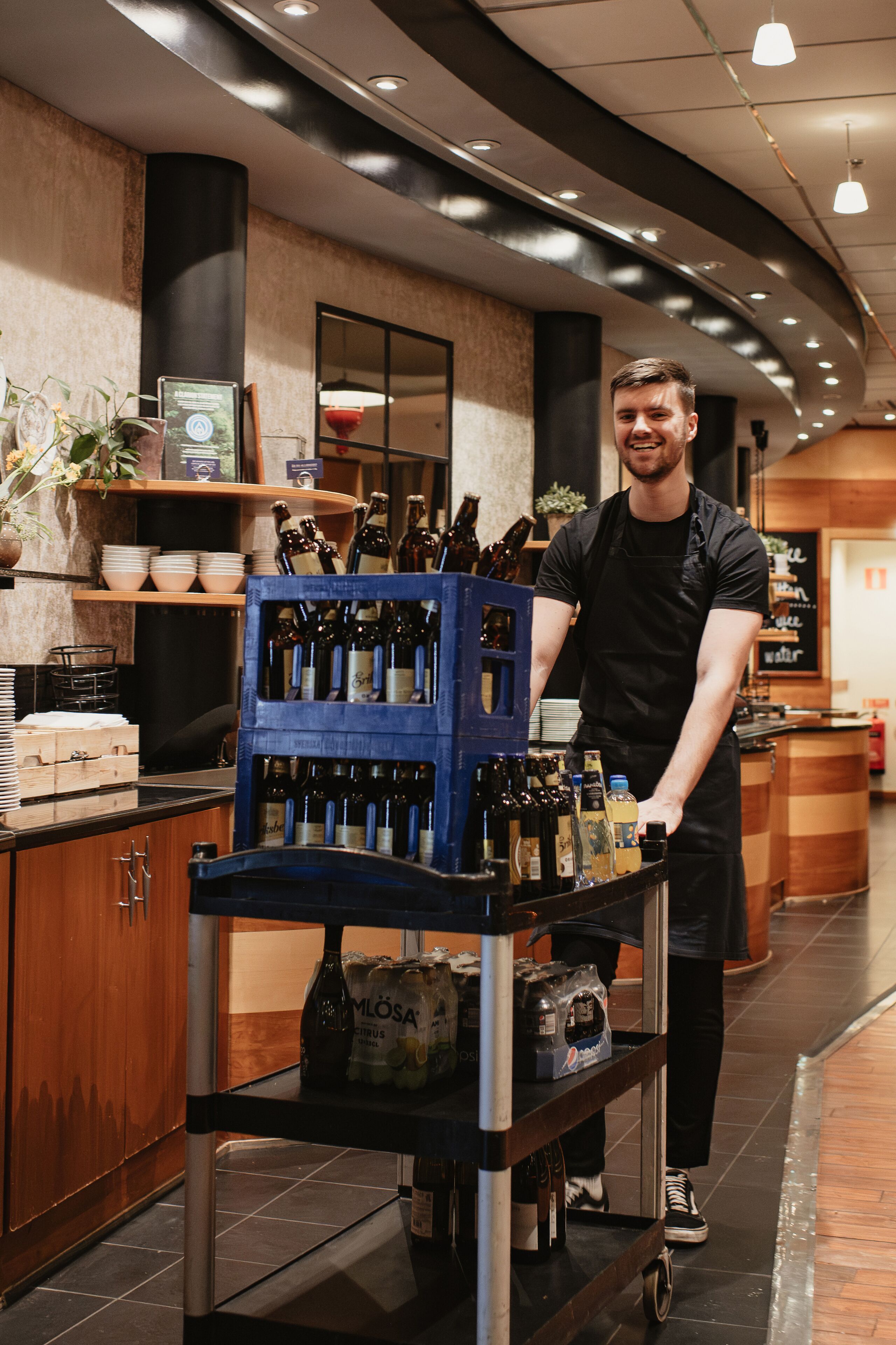 Uma área de bar de hotel com um homem ao lado de um carrinho cheio de garrafas e copos.