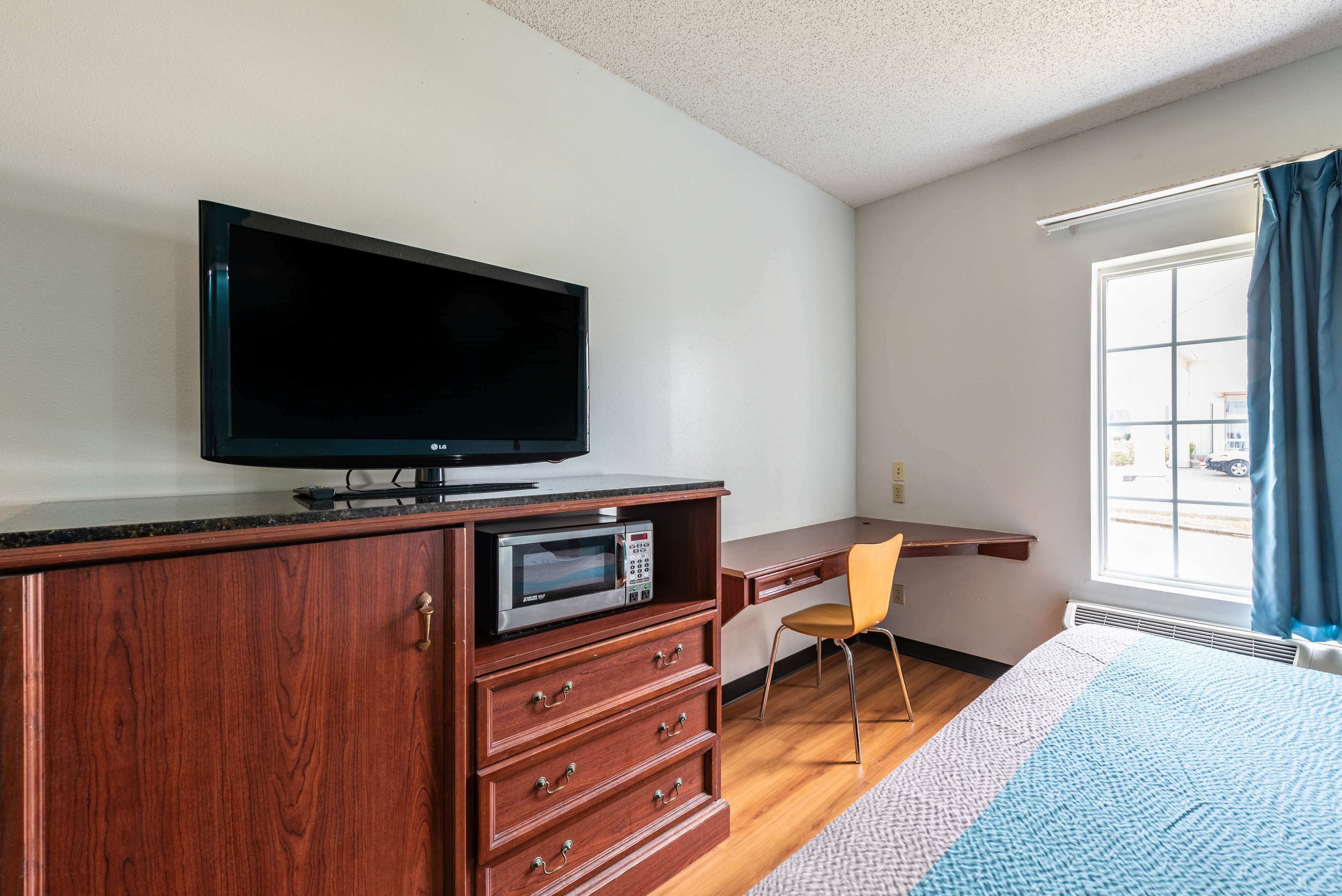 Quarto de hotel com televisão de tela plana, micro-ondas, armário de madeira, escrivaninha, cadeira e uma janela grande com cortinas.