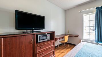 Quarto de hotel com televisão de tela plana, micro-ondas, armário de madeira, escrivaninha, cadeira e uma janela grande com cortinas.