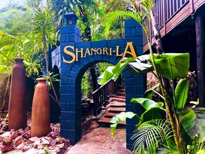 Um arco de tijolos azuis com a palavra "SHANGRI-LA" em letras douradas, conduzindo a um caminho ladeado por plantas tropicais e um piso de tijolos.