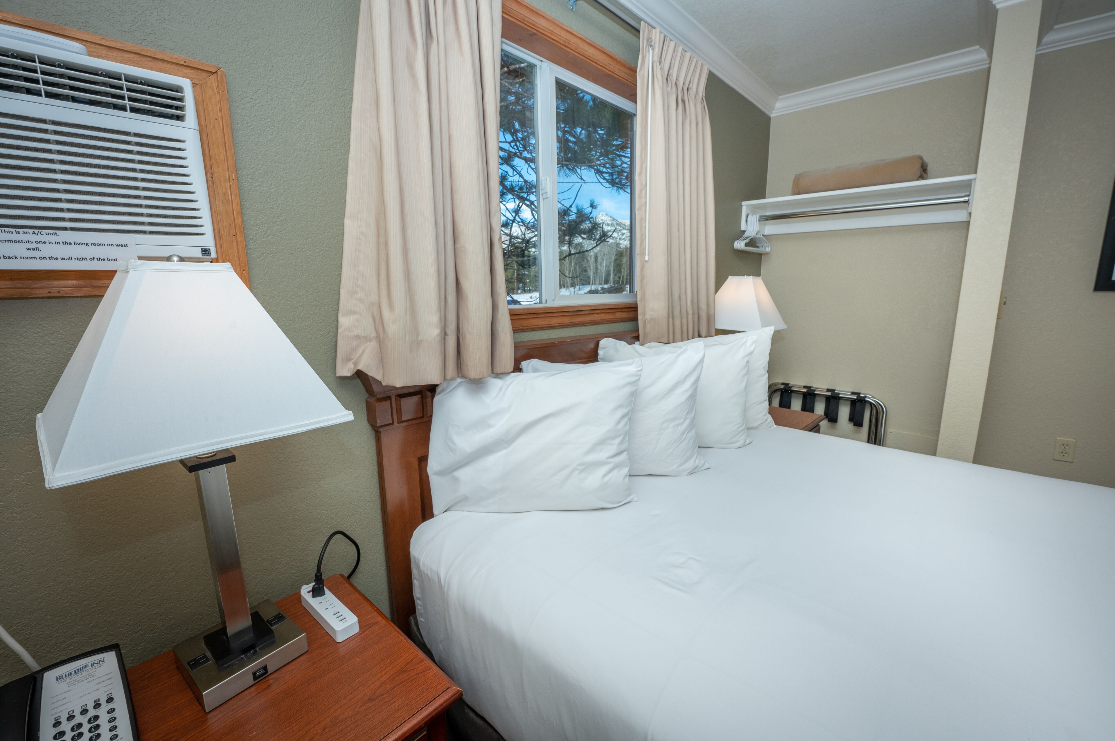 Quarto de hotel com uma cama, uma mesa de cabeceira com luminária, uma janela com cortinas e um termostato na parede.