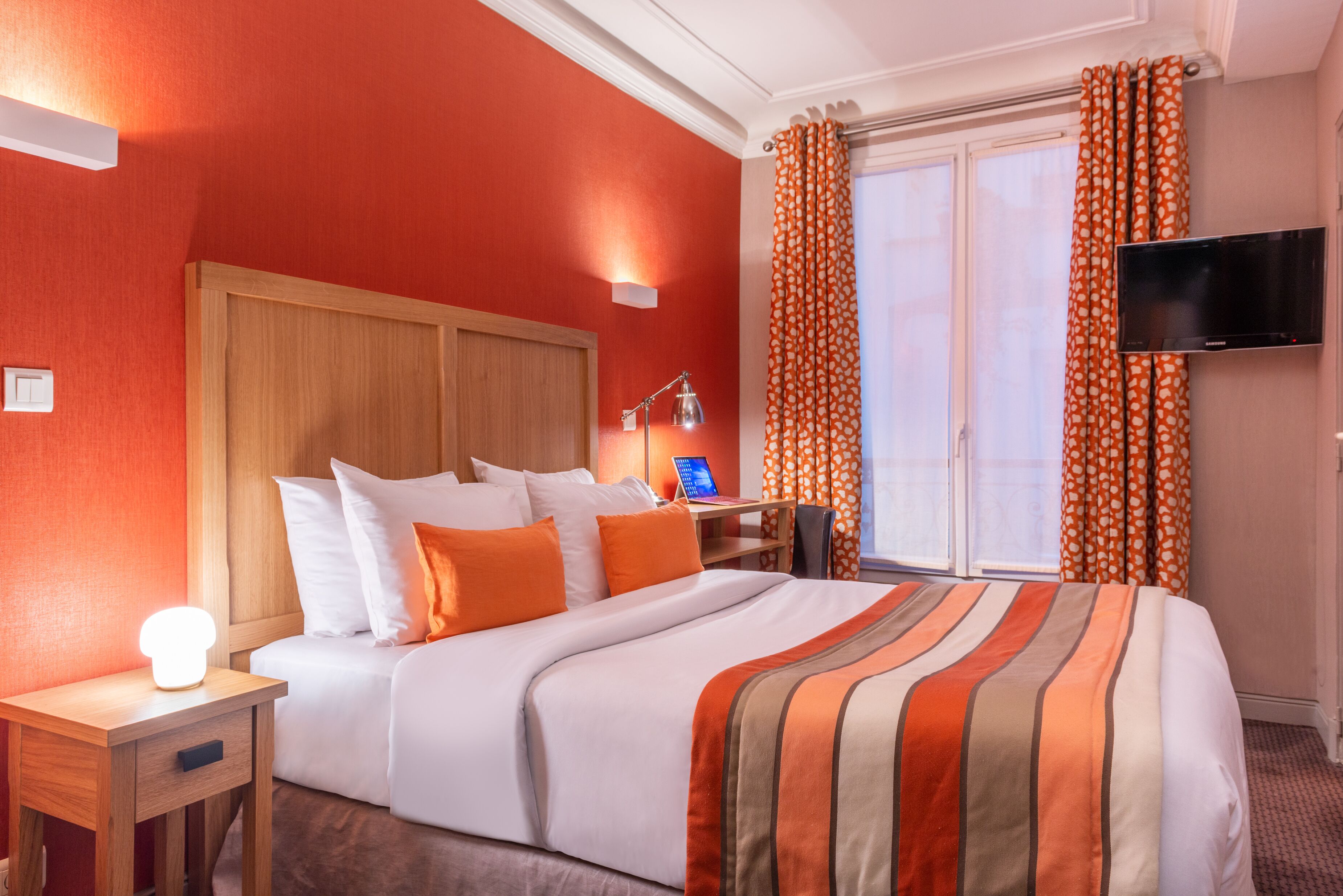 Quarto de hotel com uma cama, uma escrivaninha com candeeiro, uma televisão na parede e uma janela com cortinas.