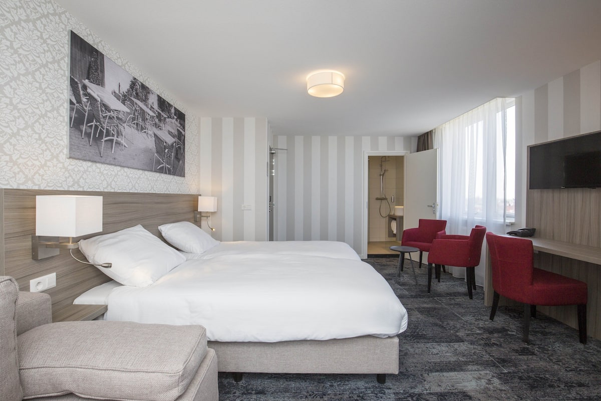 Quarto de hotel com uma cama grande, uma escrivaninha com cadeira, televisão e área de chuveiro.