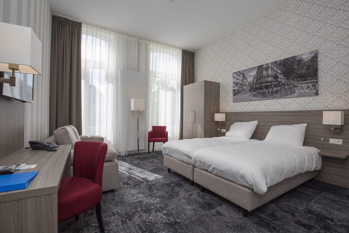 Quarto de hotel com uma cama grande, uma mesa com telefone, uma cadeira vermelha e um poltrona vermelha. Há uma janela grande com cortinas, uma televisão na parede e um quadro emoldurado.